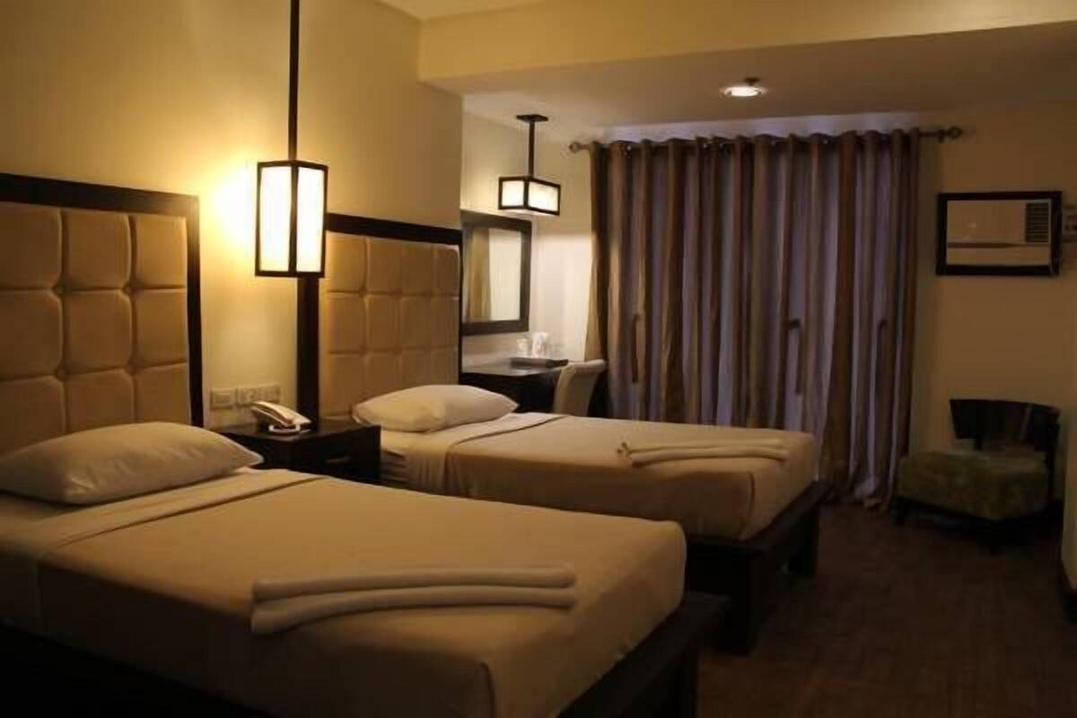 Quarto de hotel com duas camas, uma mesa de cabeceira com telefone, luminária, espelho e janela com cortinas.