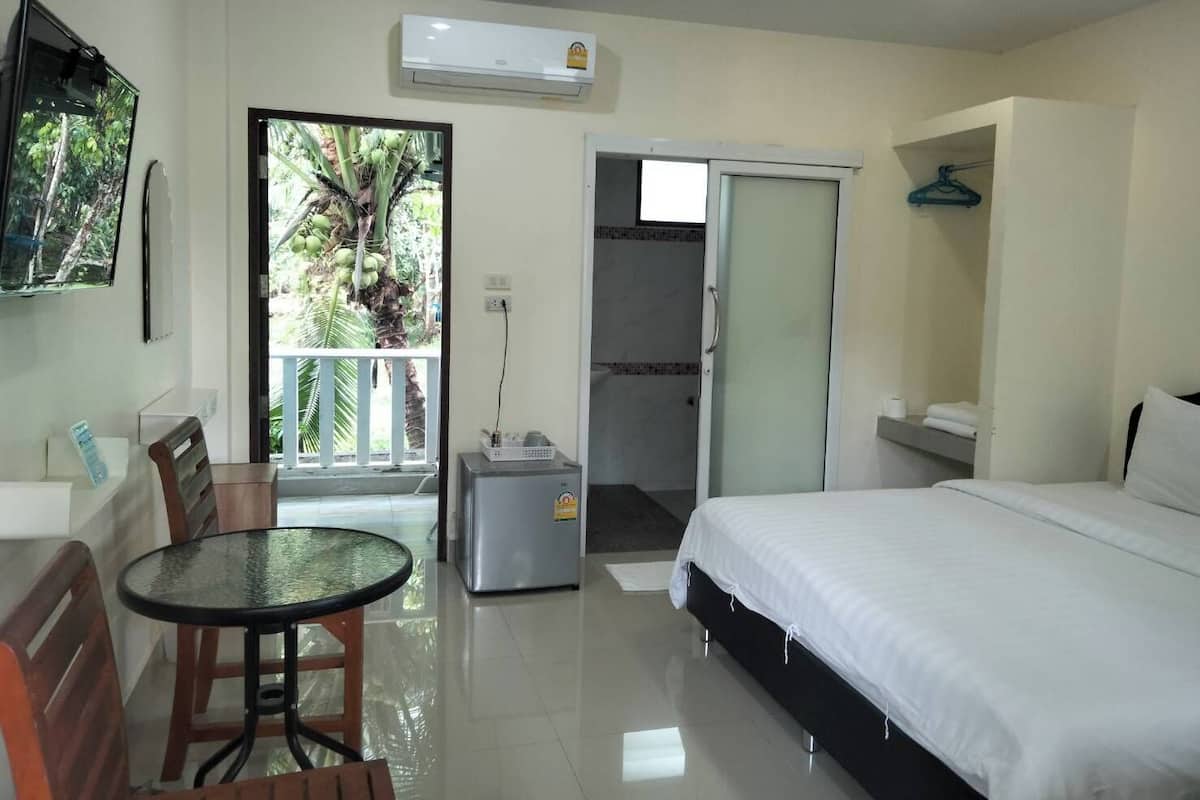 Quarto de hotel com cama, uma mesinha, uma cadeira, varanda com vista para palmeiras e porta de correr que dá para a área do chuveiro.