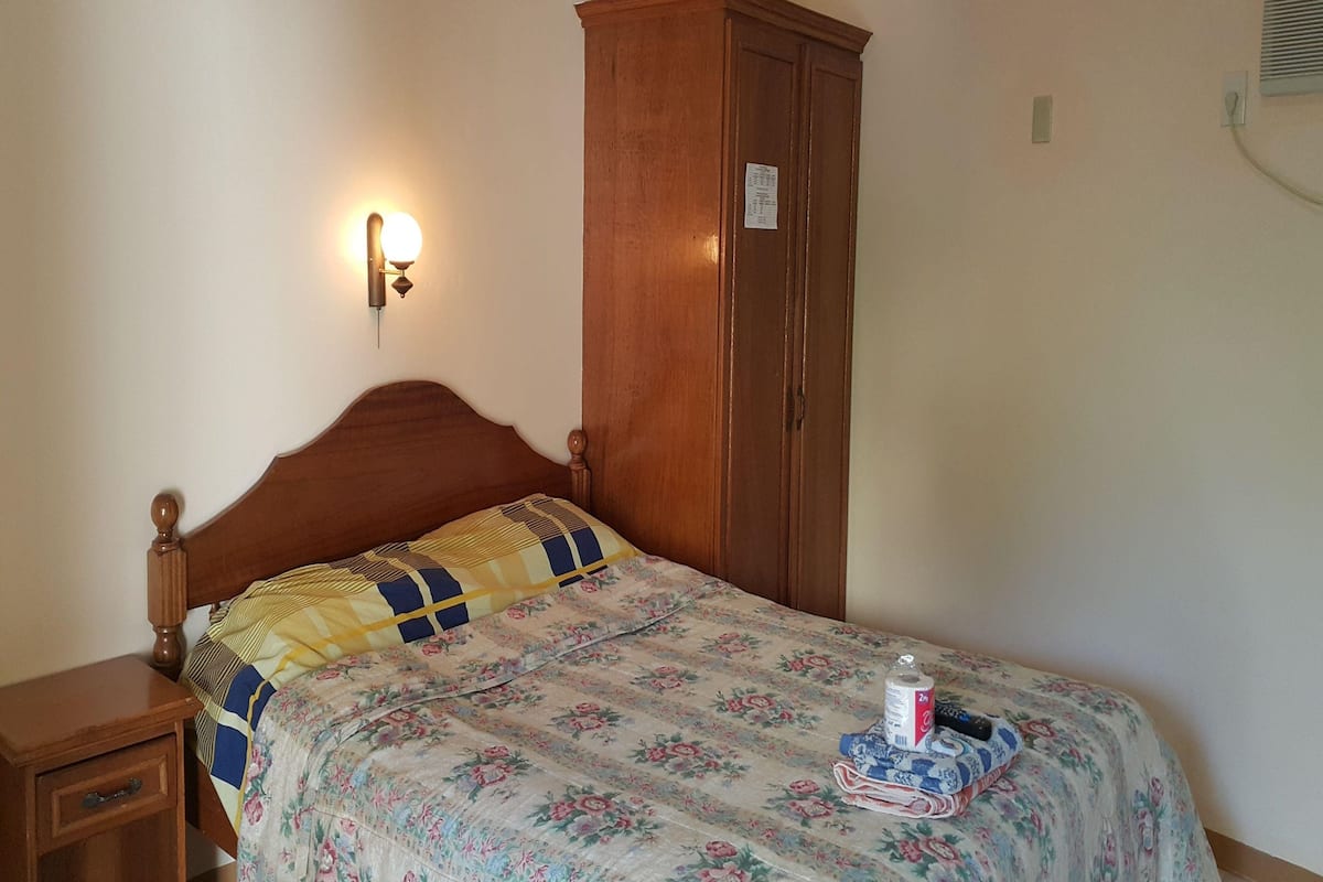 Quarto com cama de madeira, mesa de cabeceira, guarda-roupa e luminária fixada na parede.