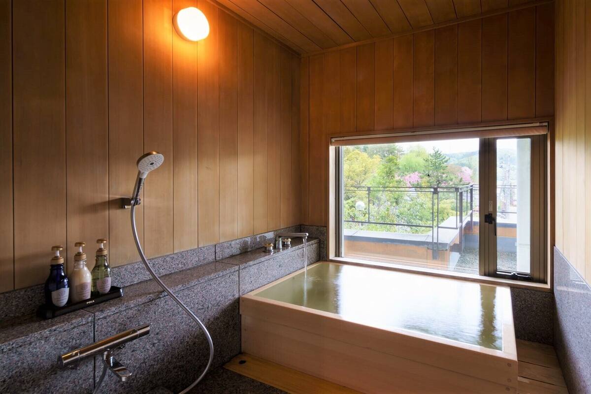 Casa de banho moderna com interior em madeira, uma banheira grande, chuveiro e janela com vista para vegetação.