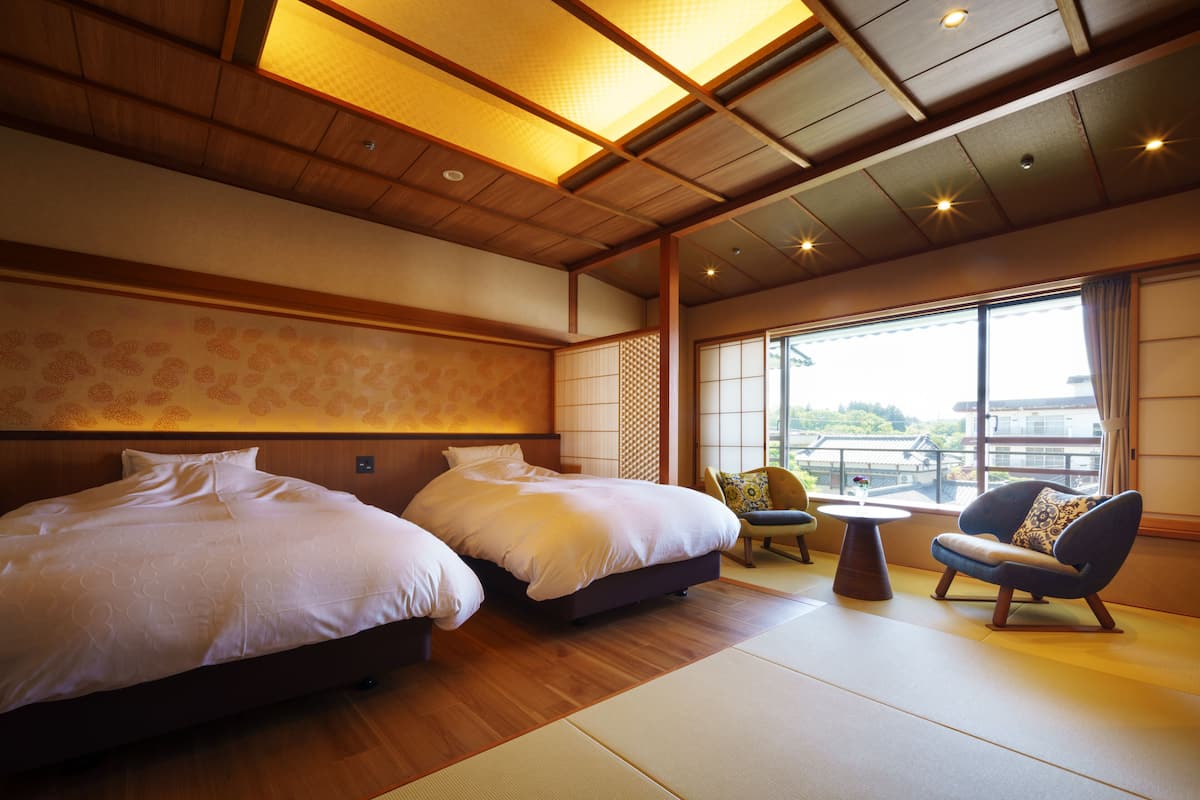 Quarto de hotel com duas camas, área de estar com uma cadeira e uma mesa pequena, e amplas janelas com vista para o exterior.