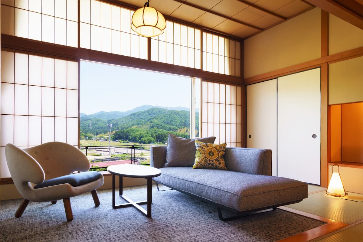 Quarto com piso de tatame, um sofá, uma cadeira e uma mesa. Uma grande janela oferece vista para uma paisagem verde.