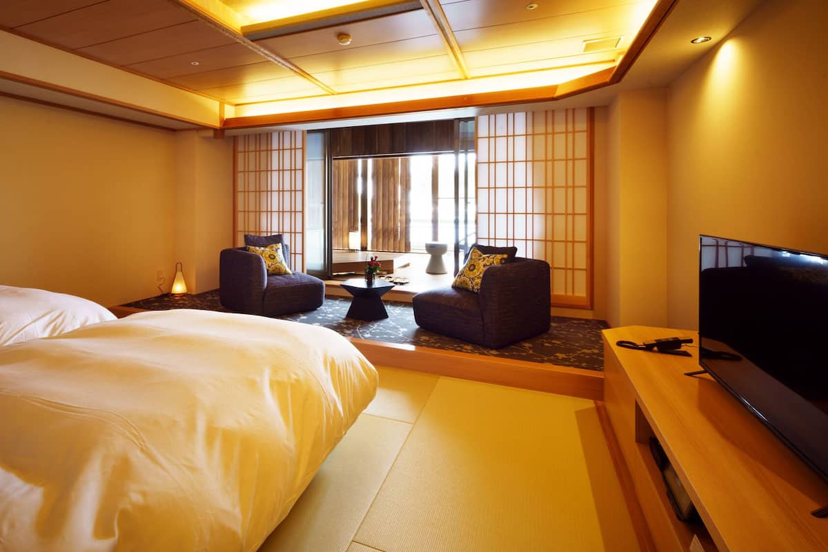 Quarto de hotel com uma cama grande, duas poltronas, uma mesa de centro, televisão e um painel deslizante shoji.