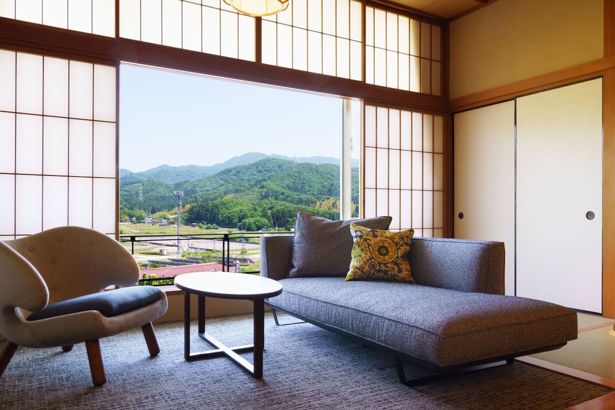 Quarto com sofá, poltrona e mesa pequena, grandes janelas com vista para colinas verdes e portas deslizantes tradicionais.