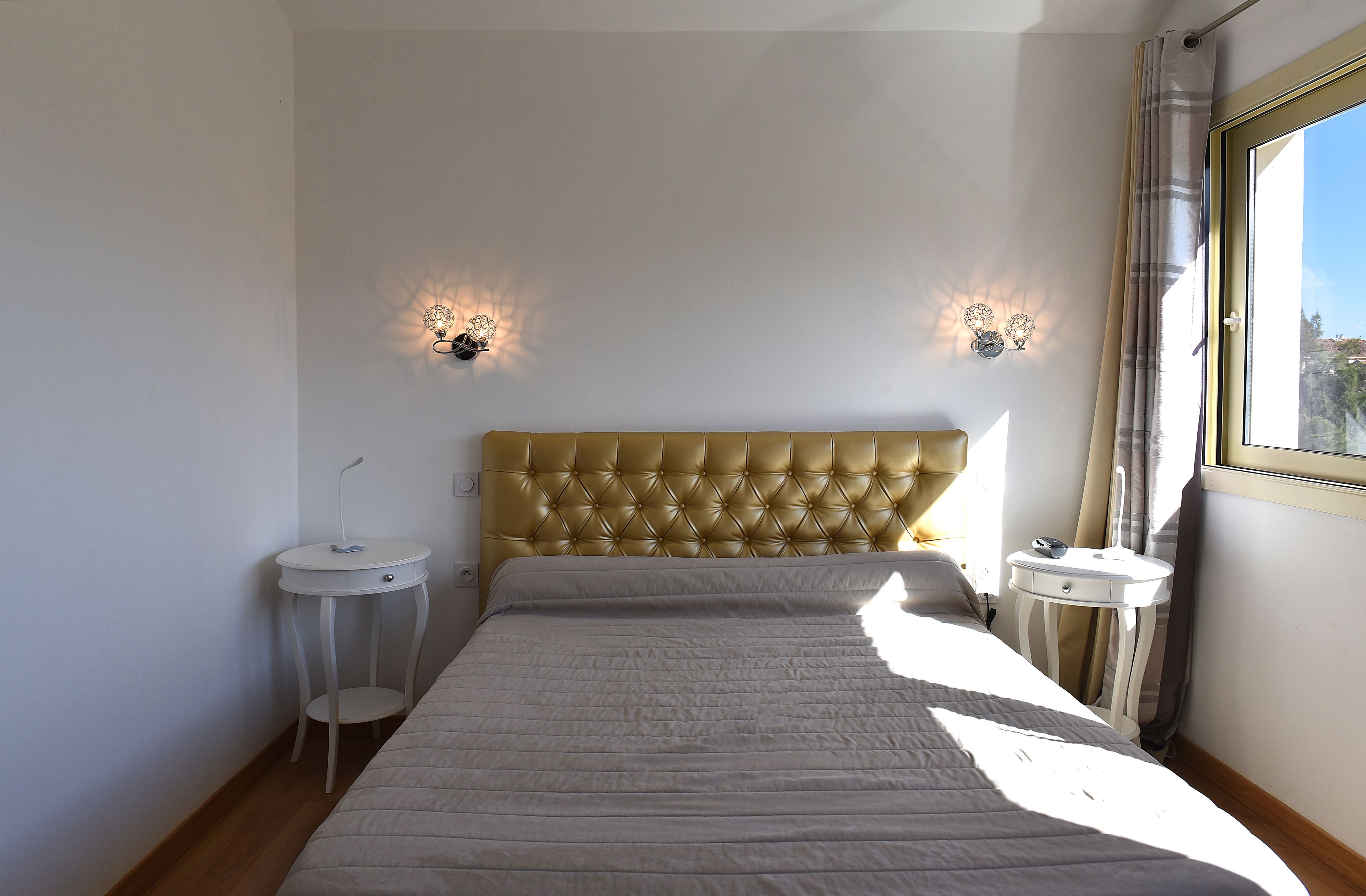 Quarto com cabeceira estofada, duas mesas de cabeceira, um criado-mudo redondo e iluminação na parede.