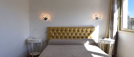 Quarto com cabeceira estofada, duas mesas de cabeceira, um criado-mudo redondo e iluminação na parede.