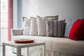 Um sofá branco com várias almofadas, uma mesinha branca com um tabuleiro vermelho e uma janela com persianas.