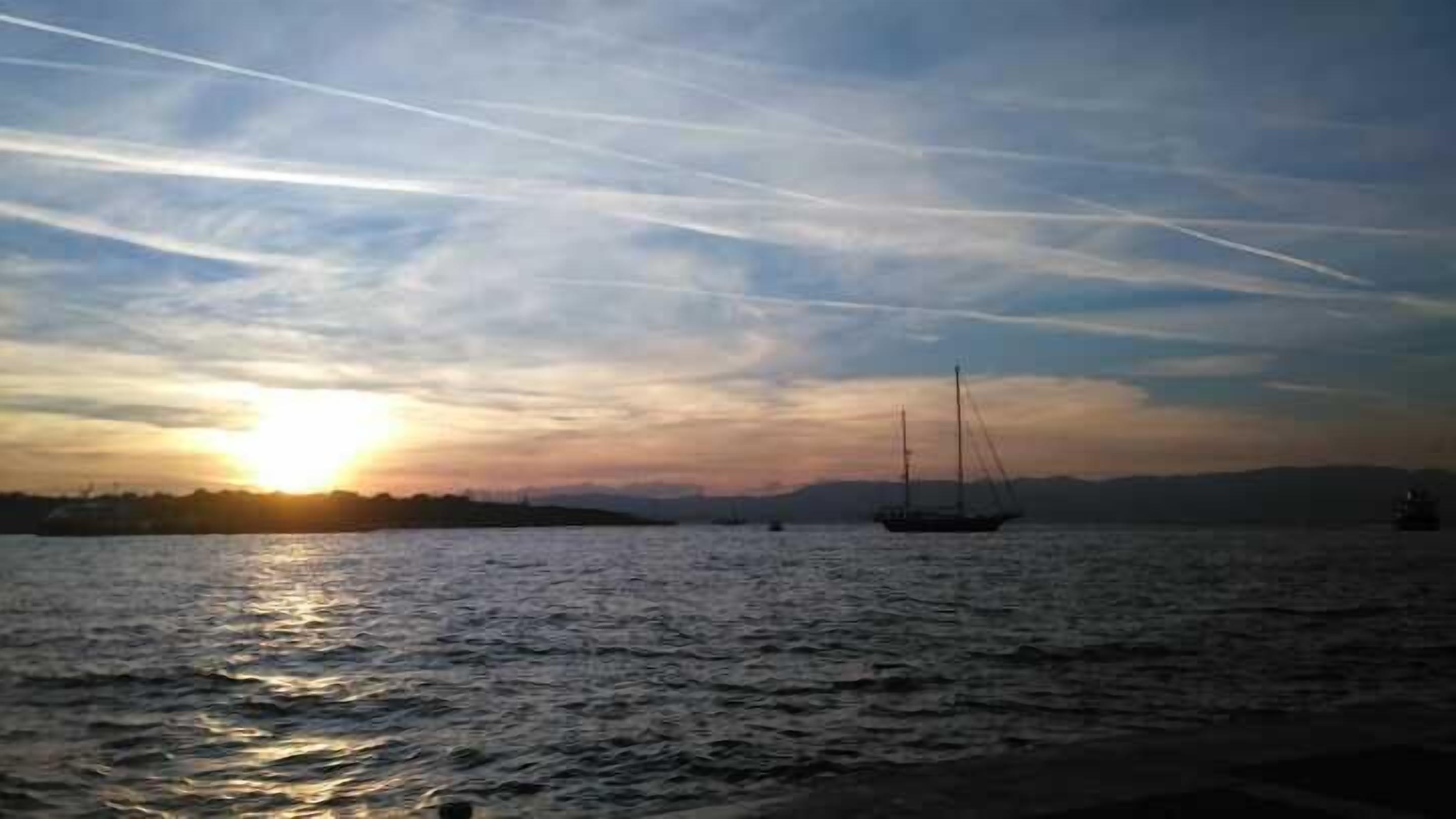 Um pôr do sol sobre águas calmas, com um veleiro e uma linha de costa distante.