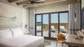Quarto de hotel com cama, escrivaninha, cadeira, ventilador de teto e vista para a varanda com móveis de exterior.