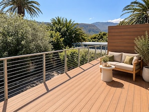 Um deck de madeira com um sofá e uma mesinha, rodeado por vegetação e vista para montanhas.