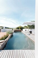 Uma piscina transparente com um deck de madeira, cercada por um muro branco e vegetação.