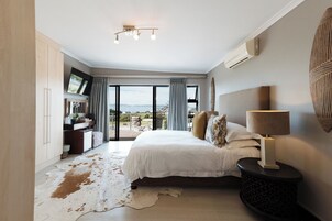 Quarto com uma cama grande, televisão fixada na parede, mesa de cabeceira com abajur e vista para o exterior através de janelas amplas.