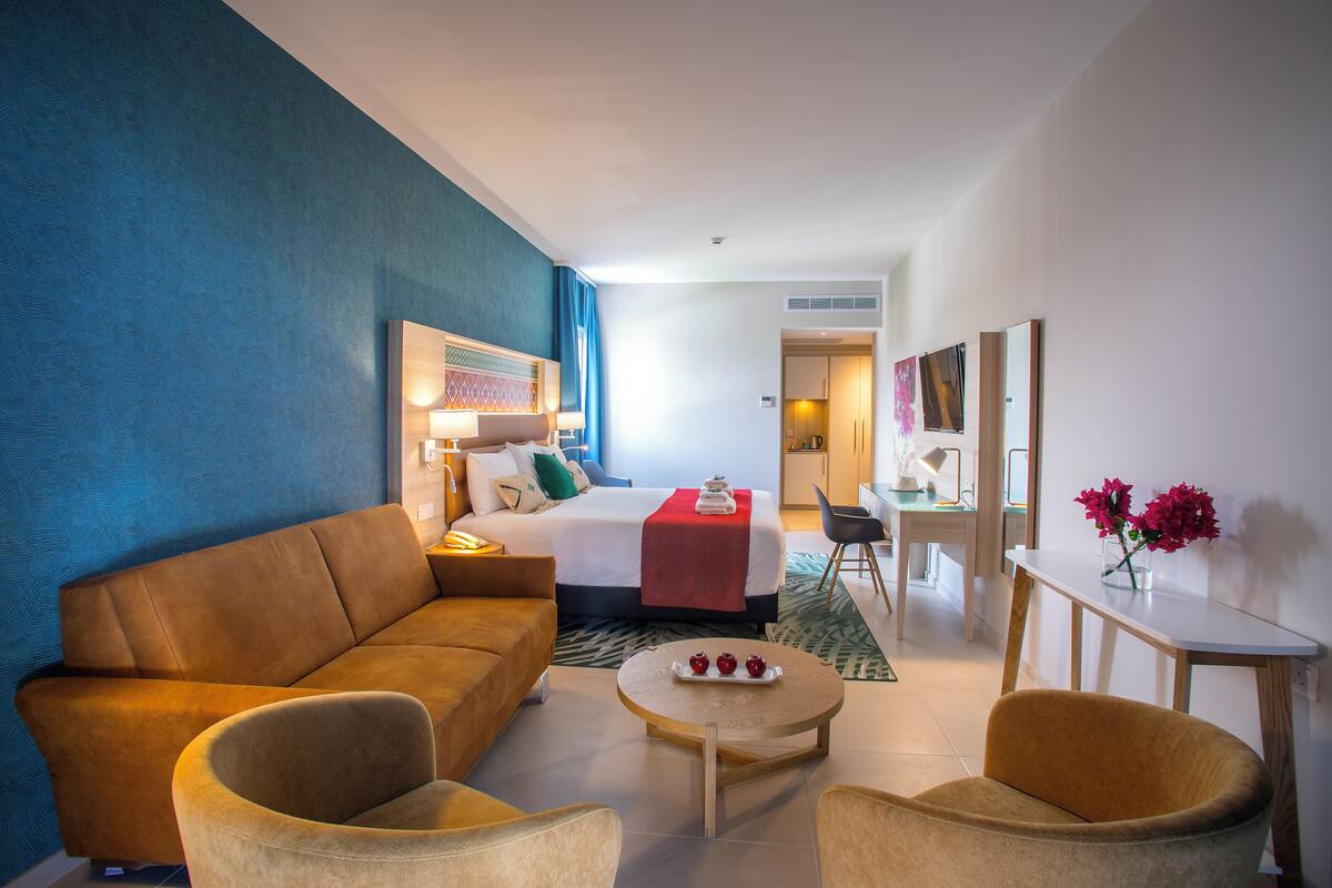 Quarto de hotel moderno com sofá, mesa de centro e cama com cobertor vermelho.