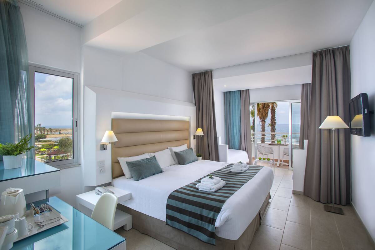 Quarto de hotel moderno com uma cama grande, uma escrivaninha com computador, uma cadeira e varanda com vista para o mar.