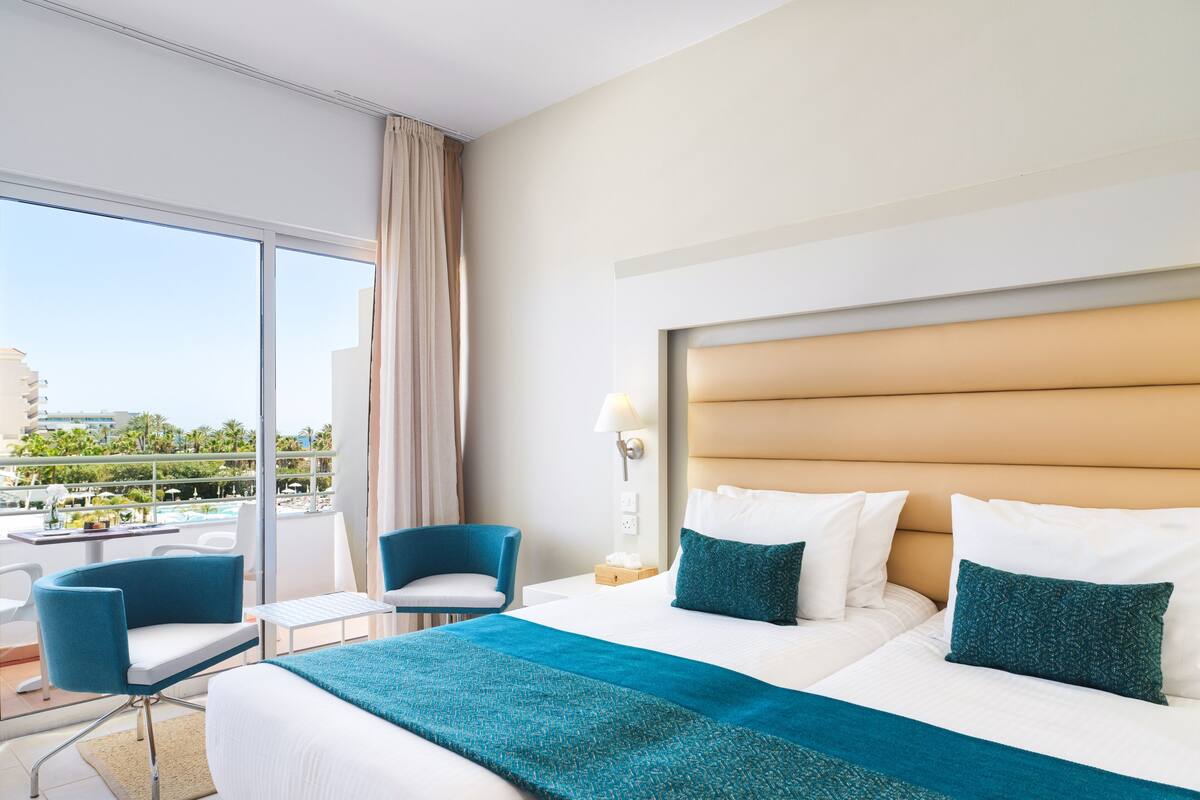 Quarto de hotel com uma cama grande, uma escrivaninha, uma cadeira e vista para a piscina e palmeiras.