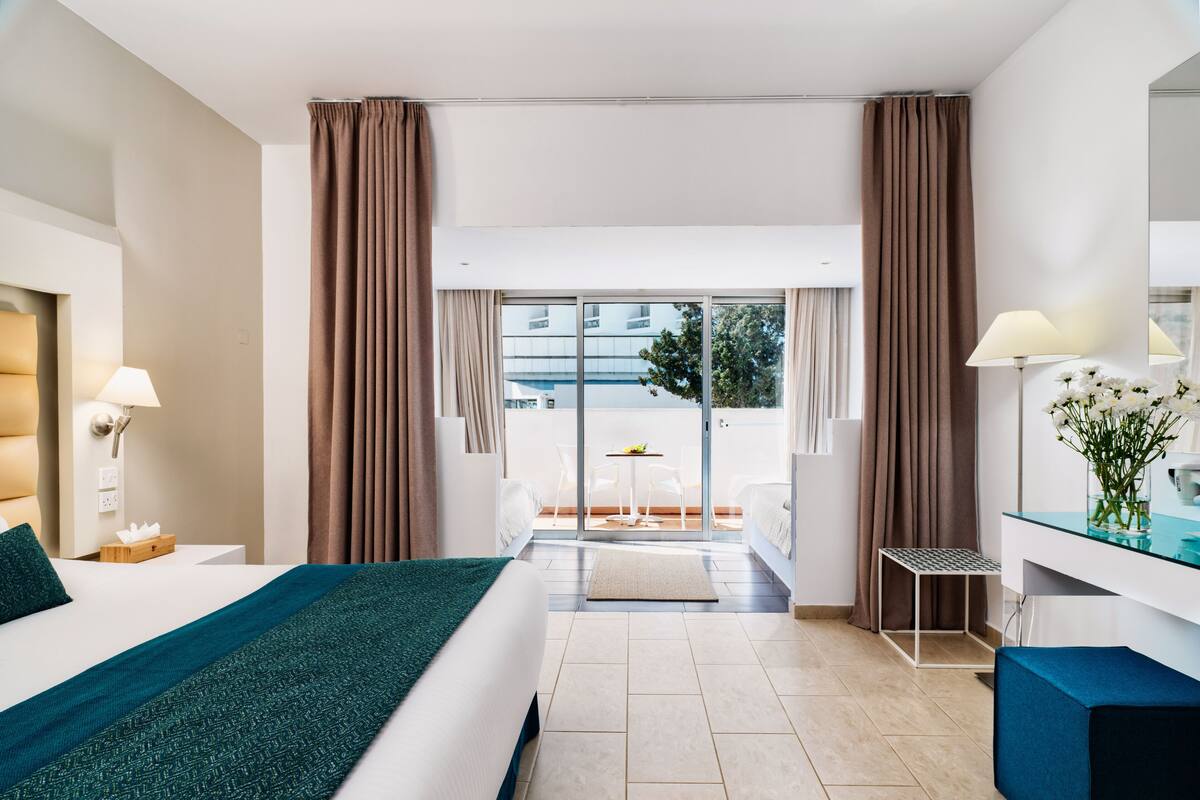 Quarto de hotel moderno com uma cama grande, uma mesa de trabalho, uma cadeira e varanda com vista para a cidade.