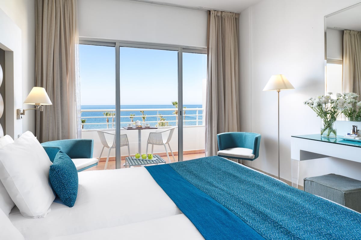 Quarto de hotel com cama grande, escrivaninha com espelho, cadeira, pequena mesa com um vaso de flores e varanda vista para o mar.