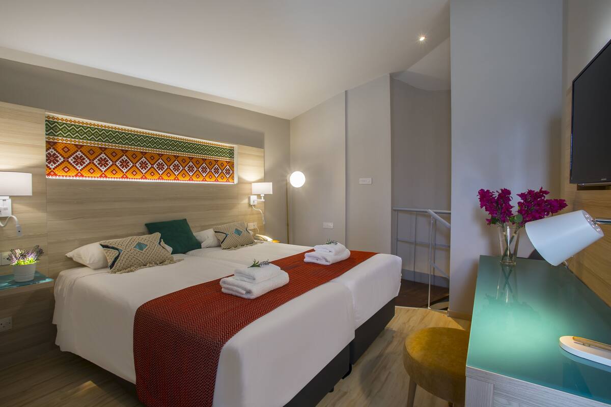 Quarto de hotel com duas camas, uma escrivaninha com um vaso de flores e uma janela grande com um padrão decorativo.