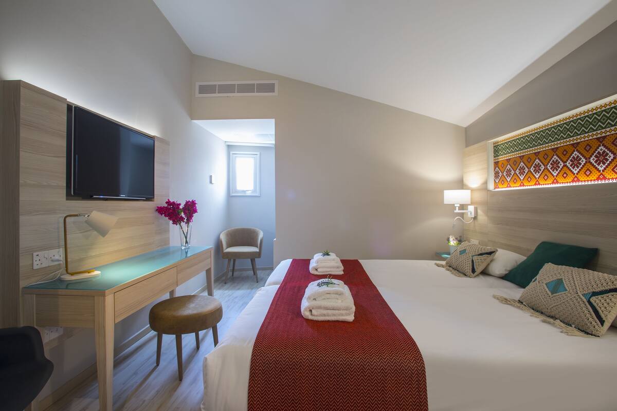 Quarto de hotel com cama, escrivaninha com computador, cadeira, televisão e janela.