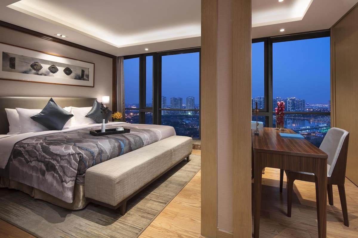 Quarto de hotel moderno com uma cama grande, uma mesa de jantar e vista para a paisagem urbana à noite.