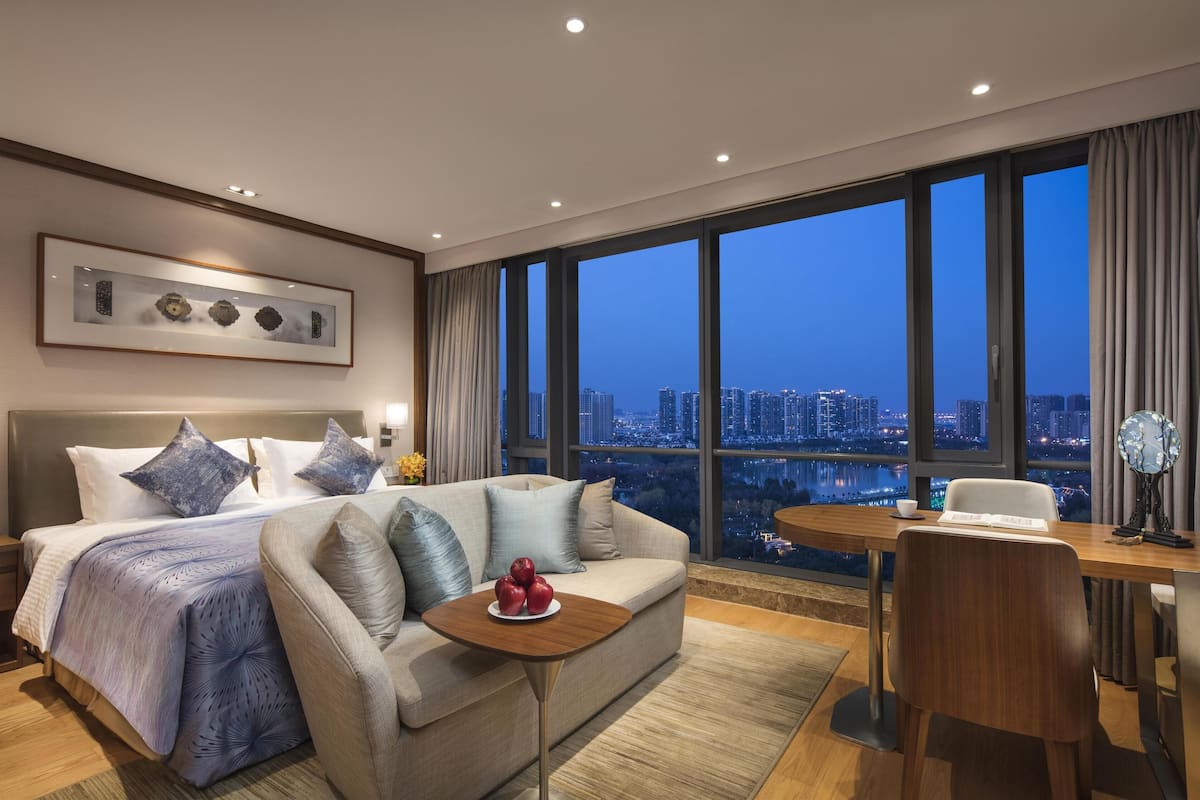 Quarto de hotel moderno com uma cama grande, um sofá, uma mesa de jantar e vista para a cidade ao entardecer.