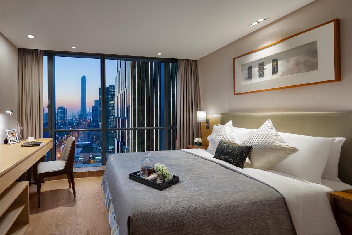 Quarto de hotel com uma cama grande, uma escrivaninha e uma cadeira. Vê-se a cidade pela janela.