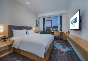 Quarto de hotel com cama, escrivaninha, cadeira e televisão na parede.