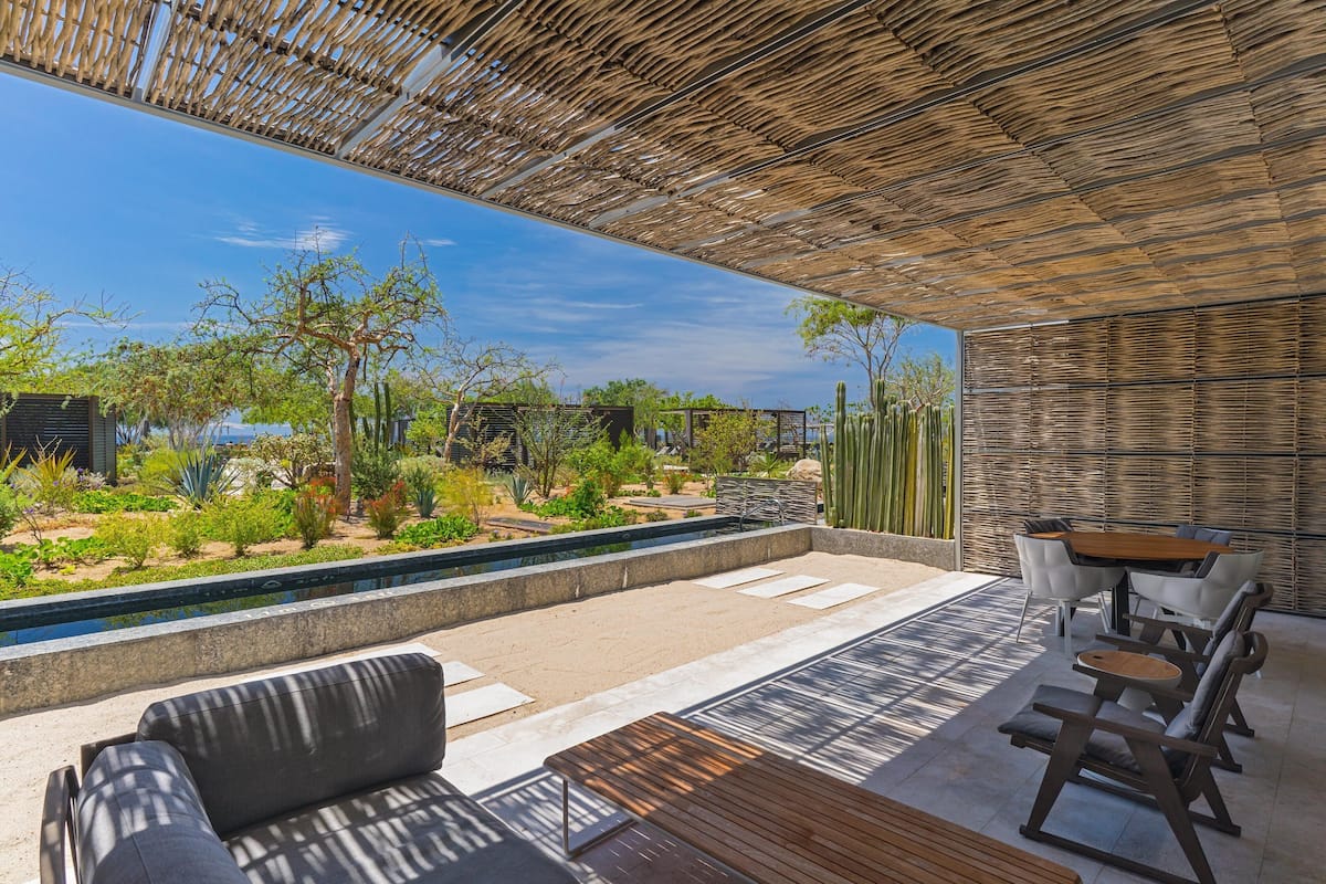 Um pátio ao ar livre com uma mesa e cadeiras de madeira, um banco acolchoado e vista para um jardim com cactos e árvores.
