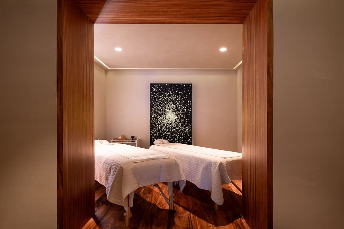 Quarto com duas camas, piso de madeira, arte de parede com tema de noite estrelada e uma pequena mesa com abajur.