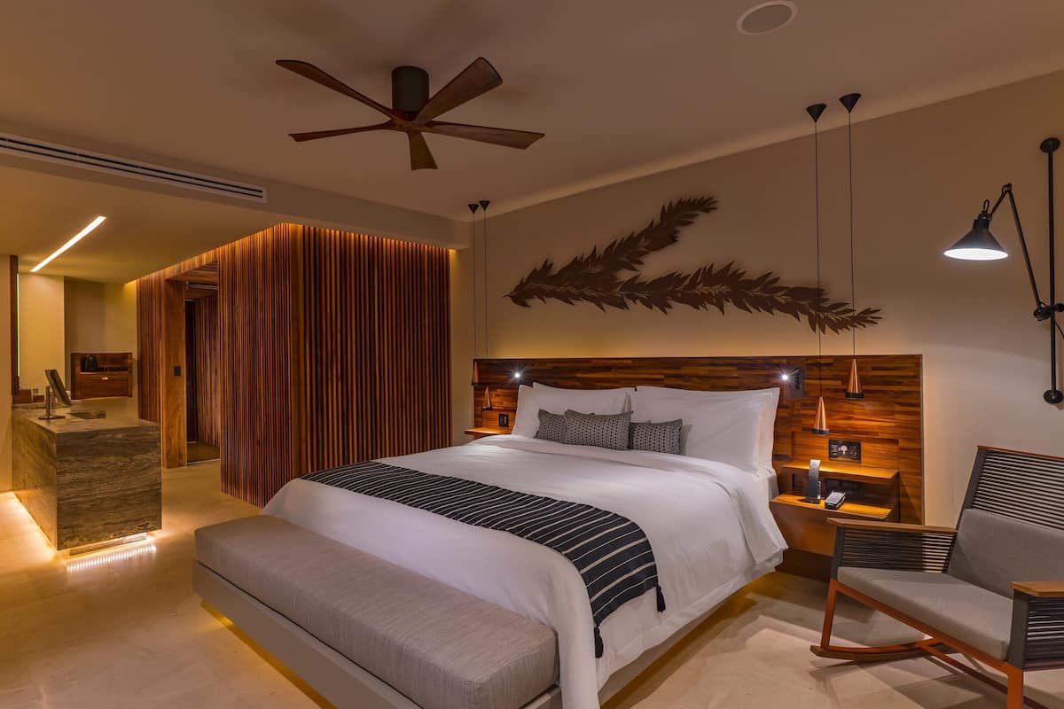 Quarto moderno com uma cama grande, mesas de cabeceira, um banco, um ventilador de teto e um detalhe decorativo na parede.
