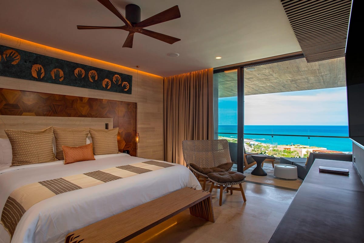 Quarto de hotel moderno com uma cama grande, ventilador de teto, vista para o mar e varanda com cadeira e mesa.