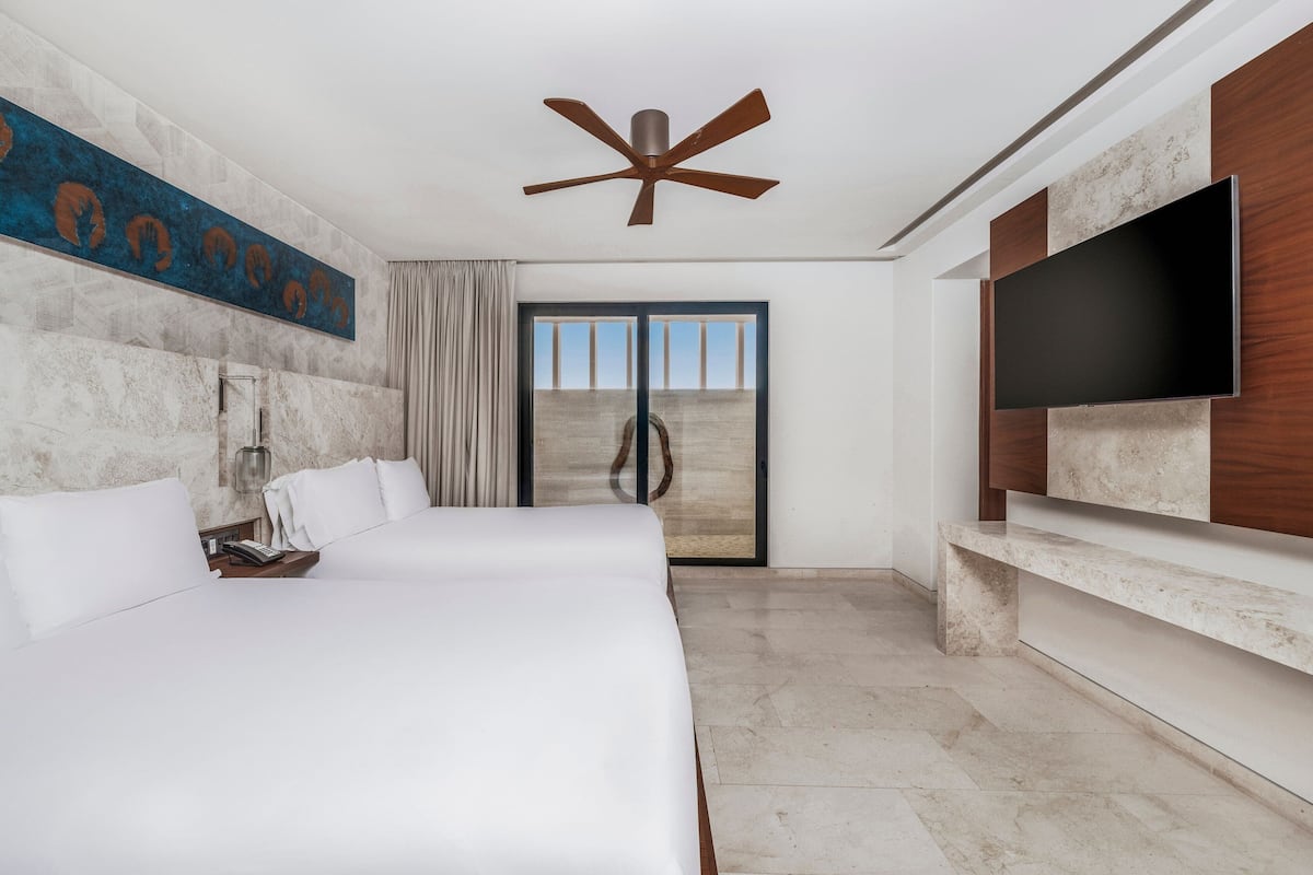 Quarto de hotel com cama grande, ventoinha no teto, TV fixada na parede e vista para o exterior por uma porta de correr.