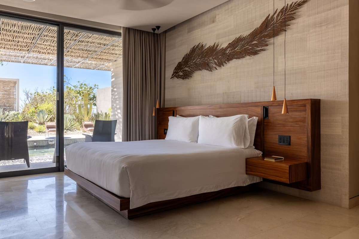 Quarto moderno com uma cama grande, cabeceira de madeira e vista para um pátio com mobiliário externo.