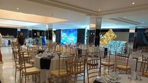 Uma sala de banquetes com mesas redondas, cadeiras de bambu e um palco com uma grande tela.