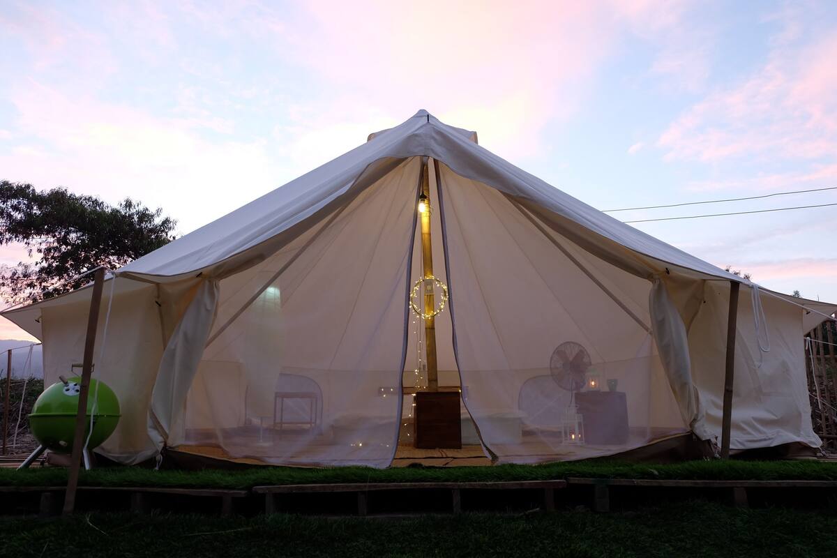 Uma tenda grande e branca com um mastro central e um candeeiro circular. No interior, há duas cadeiras e uma mesa.