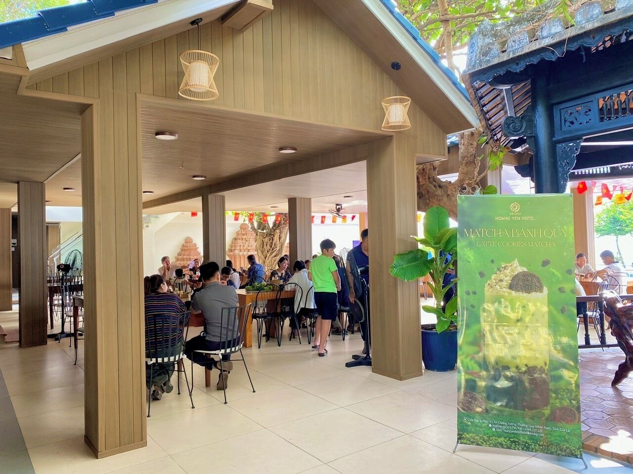 Restaurante com pilares de madeira, mesas e cadeiras, e um cartaz verde publicando um chá matcha.