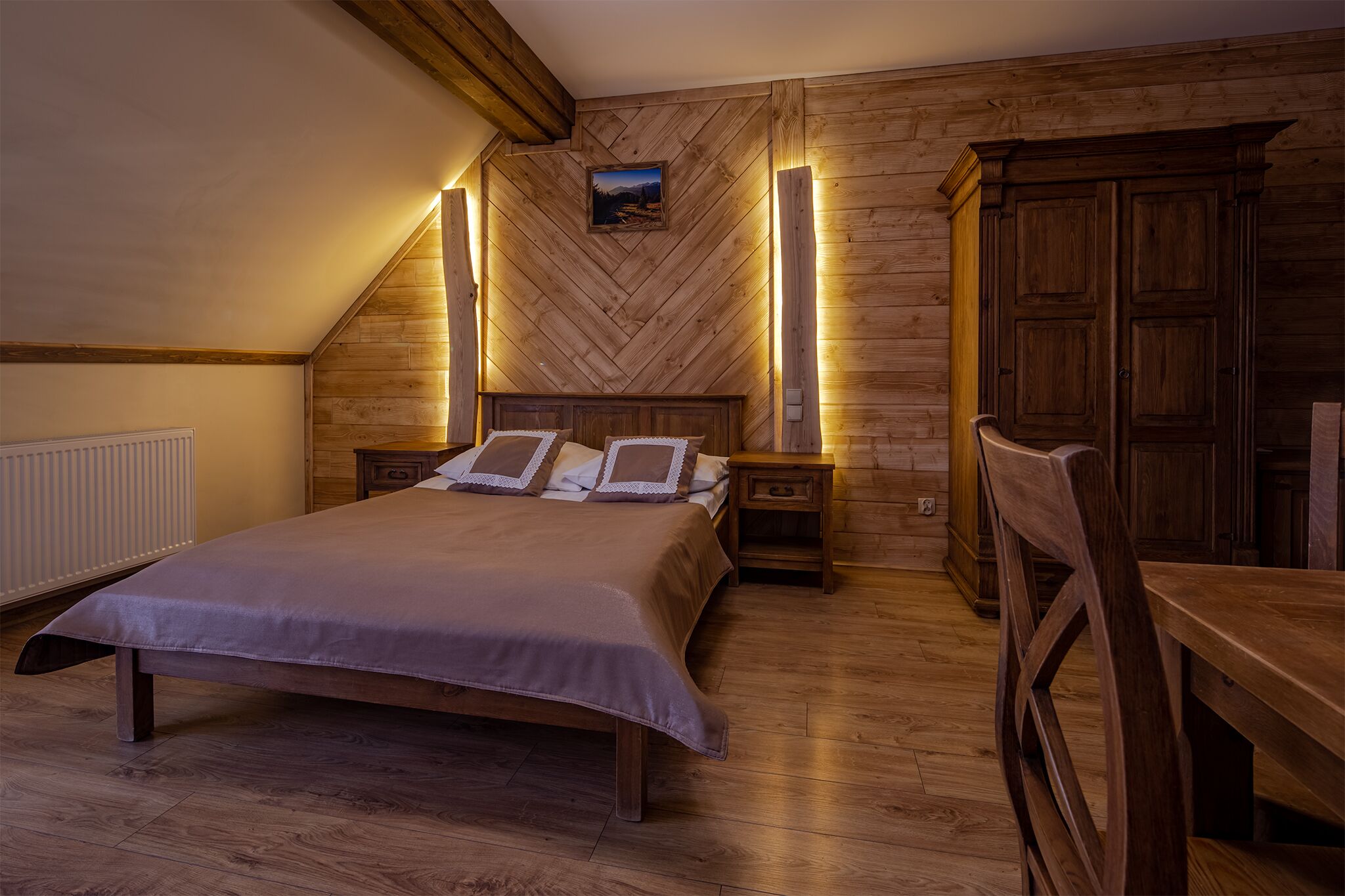 Quarto com teto de telhado inclinado em madeira, uma cama com cabeceira, mesas de cabeceira, uma cadeira de madeira e um guarda-roupa.