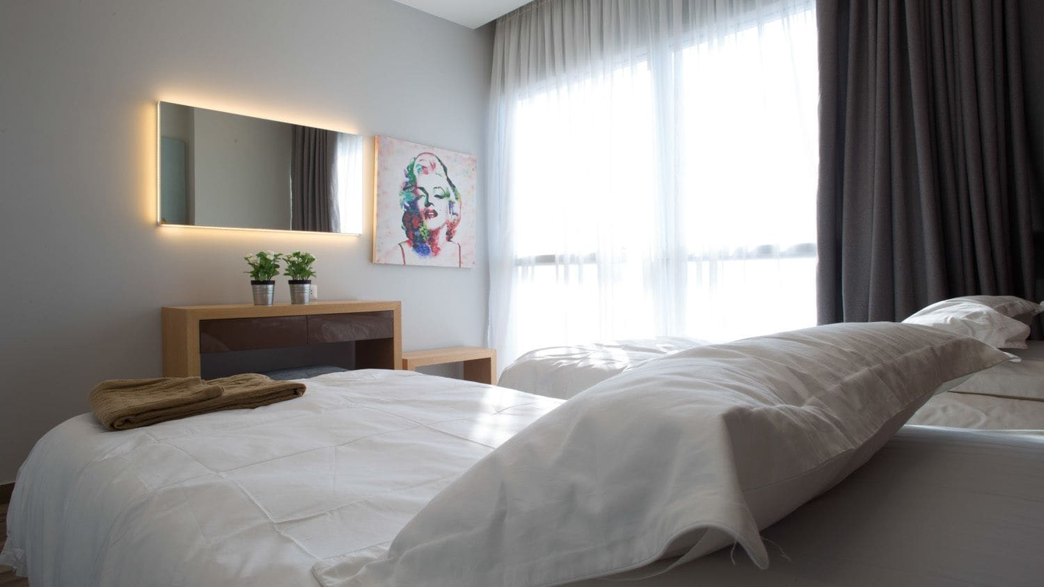 Quarto de hotel moderno com uma cama grande, uma mesa de cabeceira com abajur, um espelho e uma janela com cortinas.