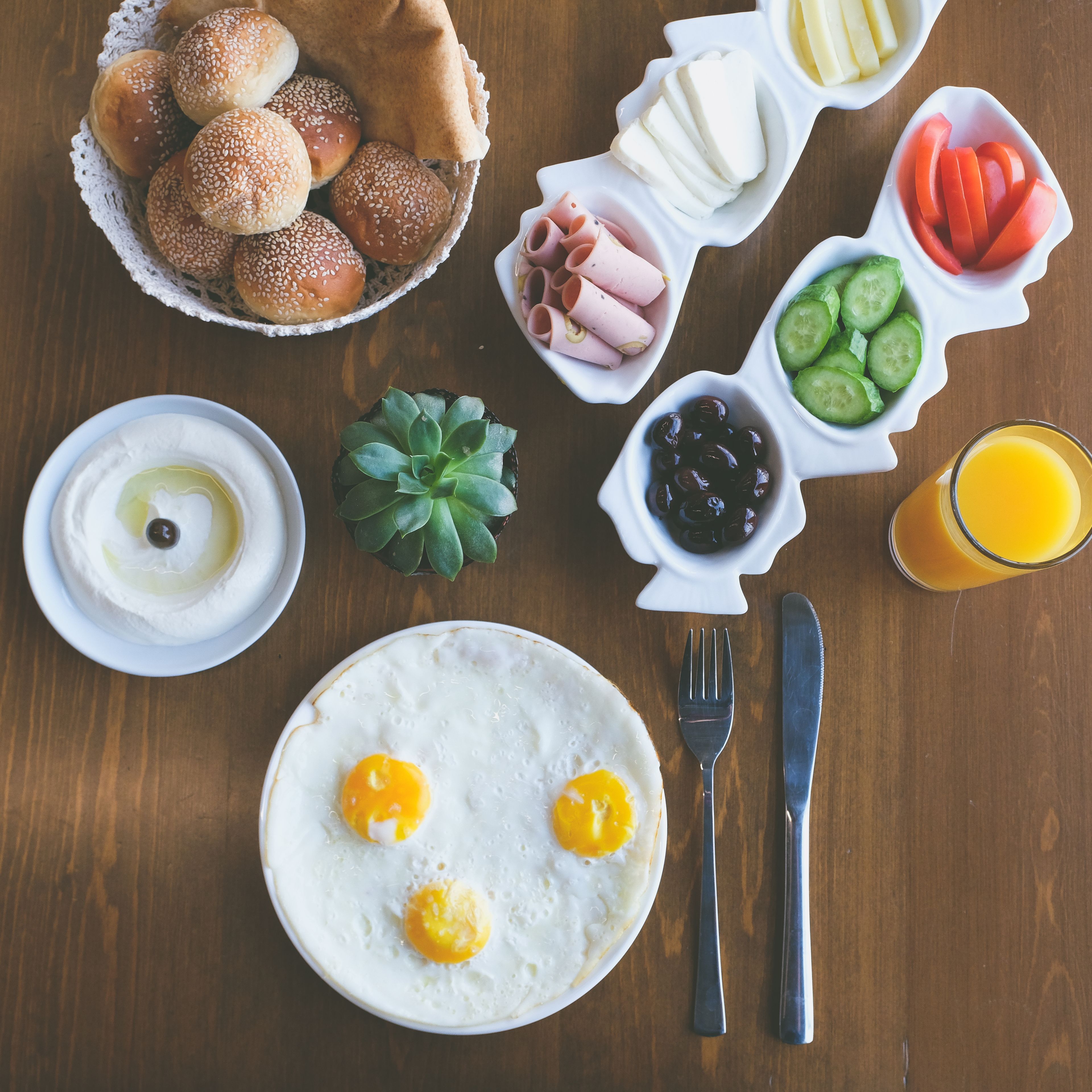 Uma mesa de café da manhã com ovos, pão, presunto, queijo, tomates, pepinos, azeitonas e um copo de suco de laranja.