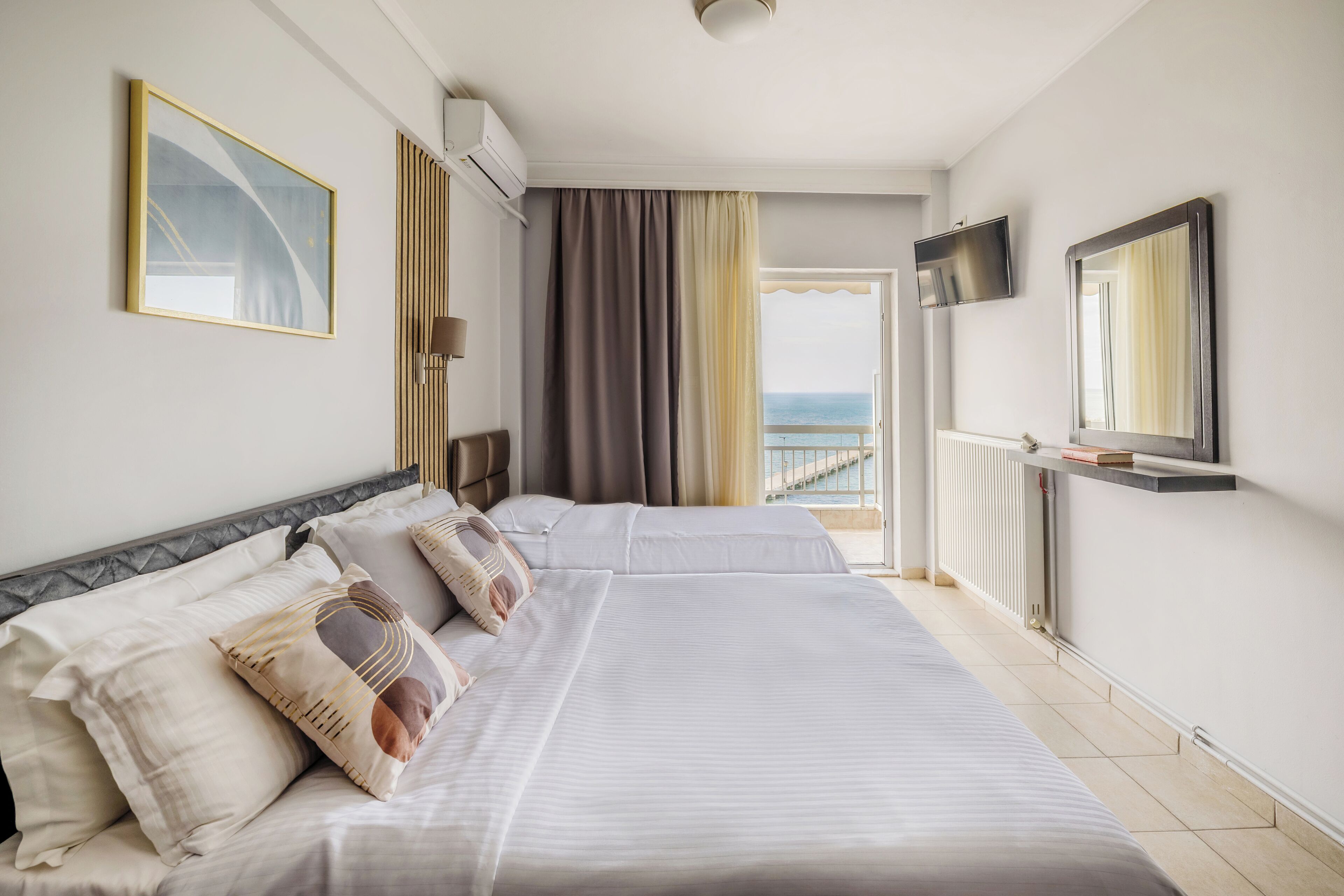 Quarto de hotel com duas camas, televisão, janela grande com cortinas e vista para o mar.