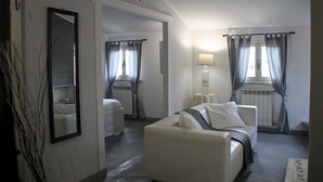 Quarto de hotel com sofá, luminária e espelho. Possui janela com cortinas e radiador.