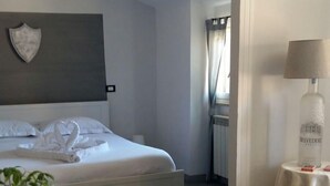 Quarto de hotel com cama, mesa de cabeceira com abajur, planta em vaso e parede com brasão.
