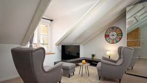 Uma sala de estar moderna com televisão de tela plana, duas poltronas cinzas, uma mesinha redonda com uma tigela de frutas e um relógio de parede colorido.