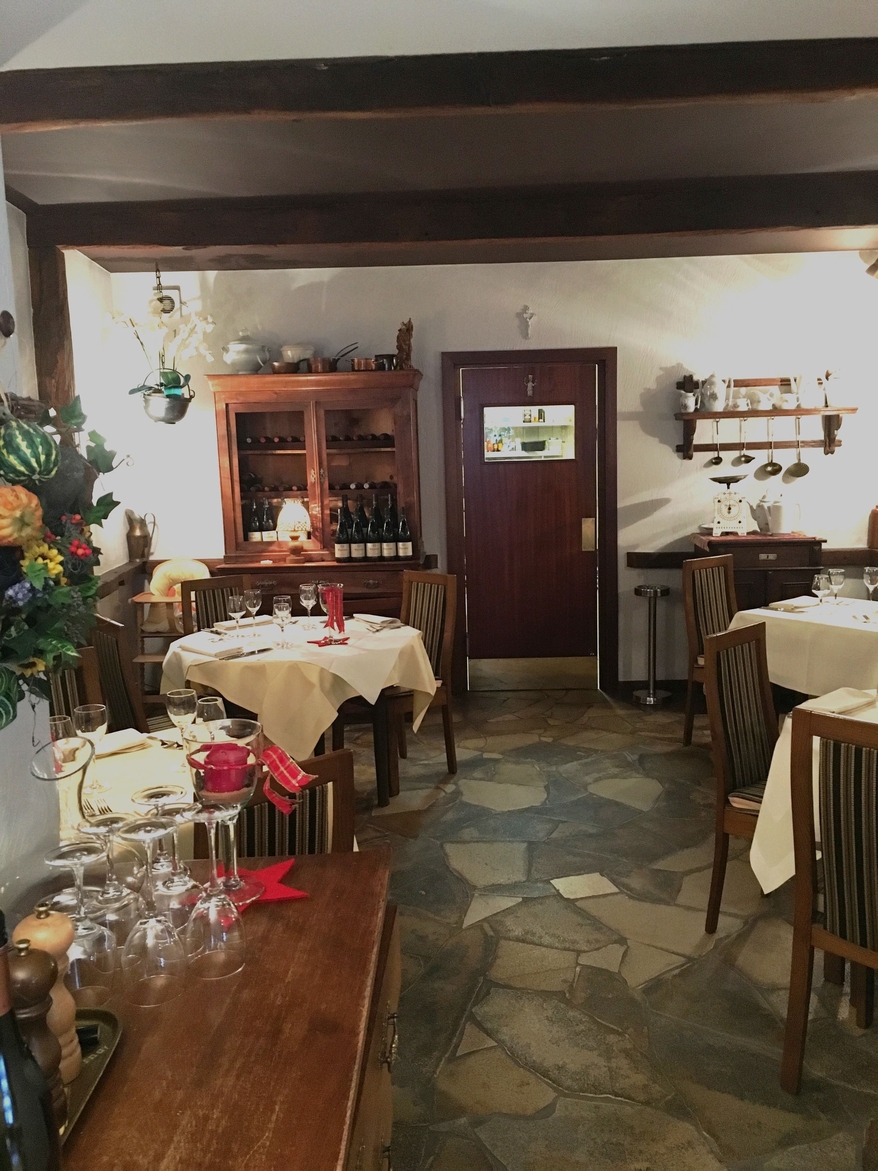 Uma área de refeições com piso de pedra, mesas e cadeiras de madeira. Há garrafas de vinho e copos sobre as mesas, e um armário de madeira com prateleiras.