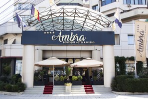Exterior do Ambra Boutique Hotel & Bistro com design arquitetônico moderno, destacando uma entrada imponente com degraus e duas grandes ombrelhas oferecendo sombra.