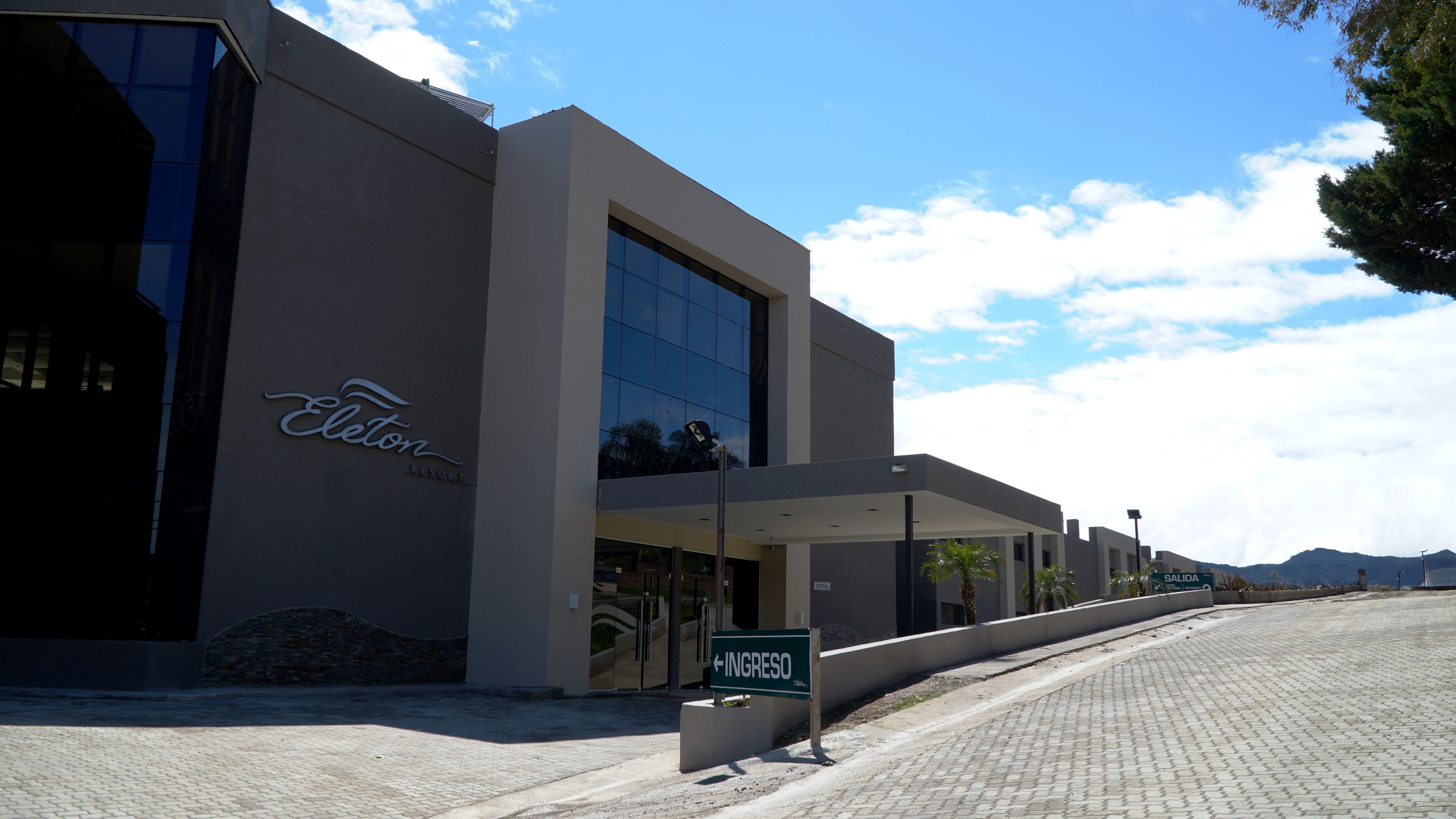 Um edifício moderno com uma fachada de vidro ampla, uma placa indicando 'Ingresso' para a entrada e uma placa com 'Saída' para a saída.