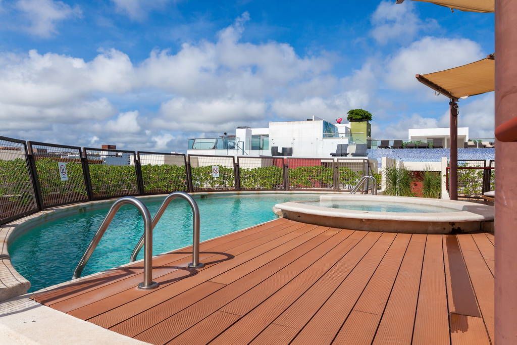 Uma área de piscina com deck de madeira, uma banheira de hidromassagem e vista para edif ícios ao fundo.
