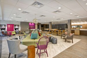 Um lobby espaçoso com diversos espaços de estar, incluindo um sofá verde, uma poltrona roxa e várias poltronas. Há várias mesas e cadeiras dispostas para refeições ou convívio. Uma placa em destaque diz "HOME2".
