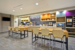 Um restaurante moderno com várias mesas e cadeiras, um balcão com cardápio e uma tela de televisão.
