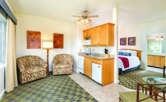 Quarto de hotel compacto com cozinha compacta, duas poltronas estampadas, cama com manta colorida e obras de arte nas paredes.